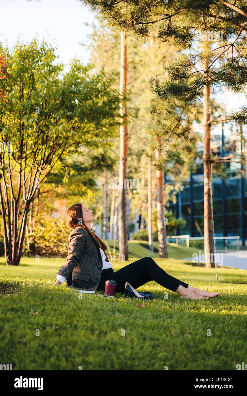 Full length of a relaxed businesswomen sitting on green grass enjoying ...
