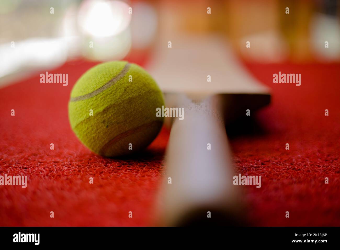 A closeup shot of a cricket bat and a ball on a red surface with a blur ...