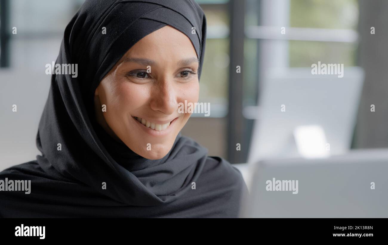 Cute positive arab woman in hijab looks at laptop screen smiling ...