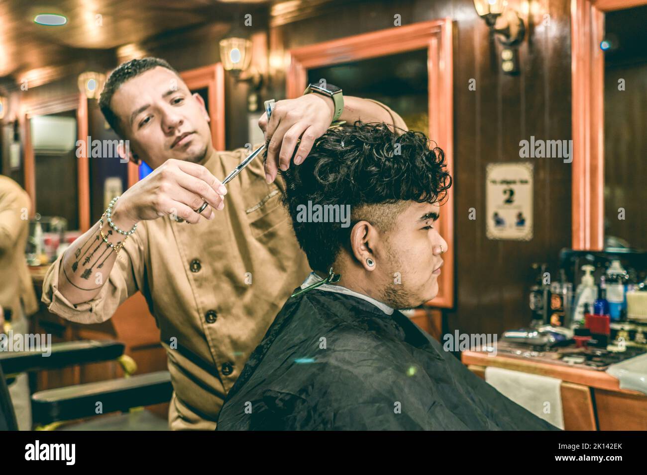 Serious tattooed barber cutting hair of young man with piercings ...