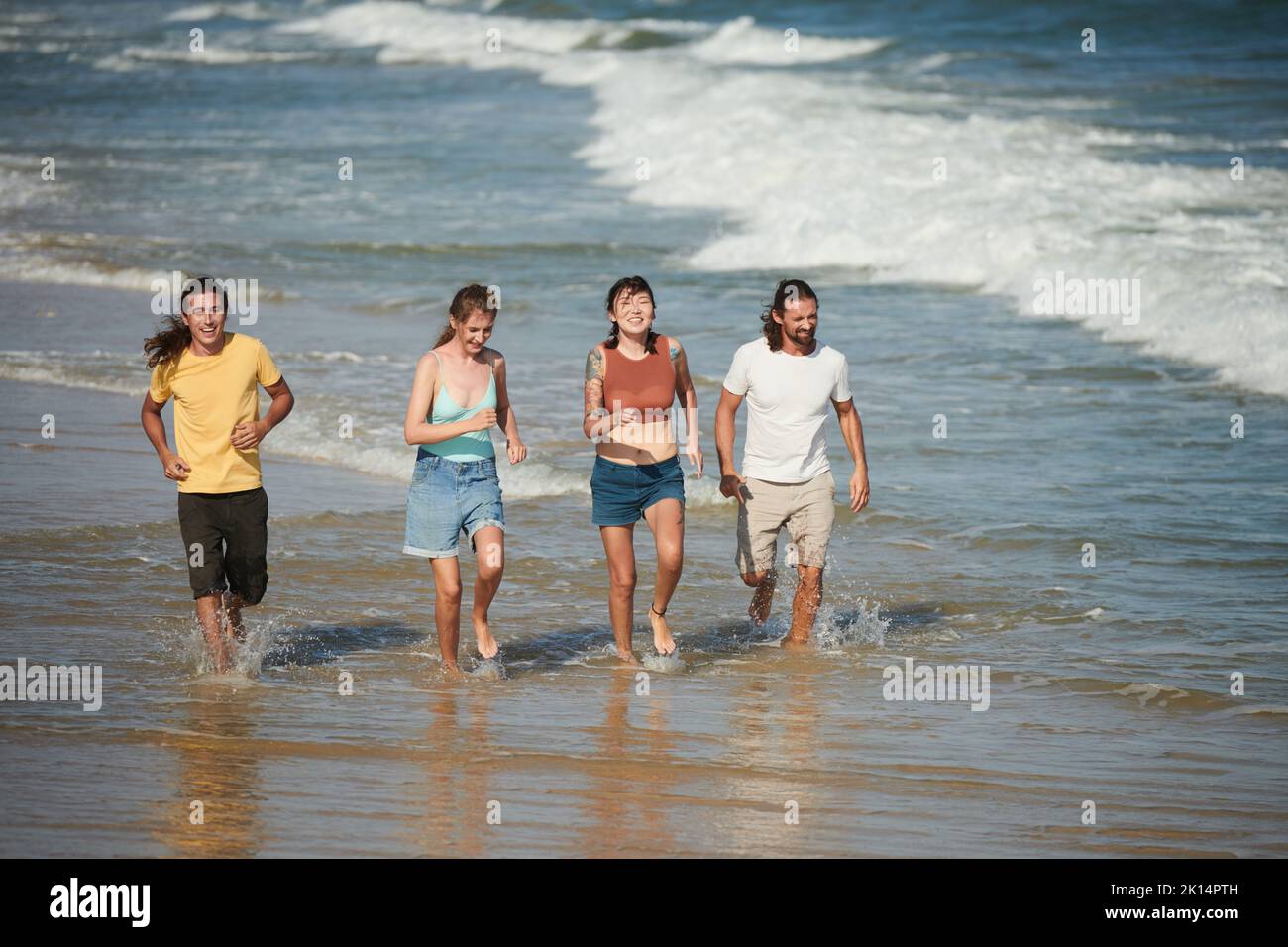 Joyful young people running on beach, splashing water and laughing ...
