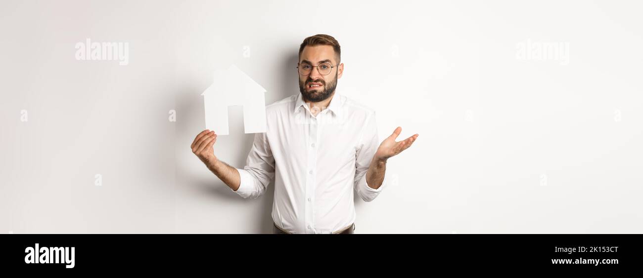 Real estate. Confused man shrugging, showing house paper model and ...