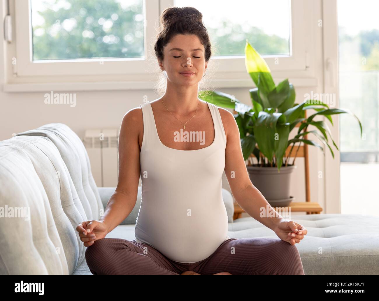 Pregnant woman sitting in lotus position and meditating on sofa in ...