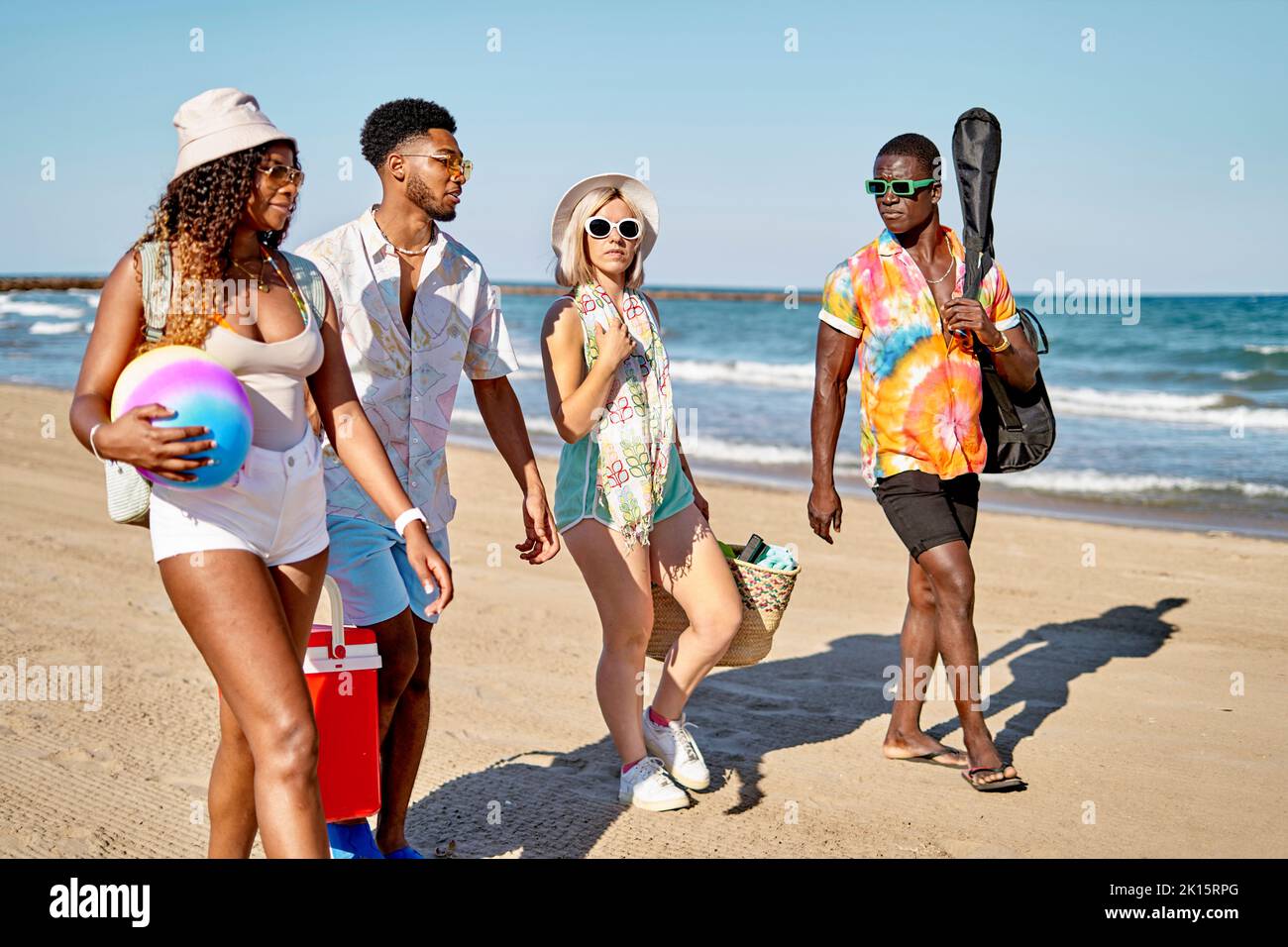 Full body multiracial men and women in sunglasses and hat strolling on sand against cloudless ...