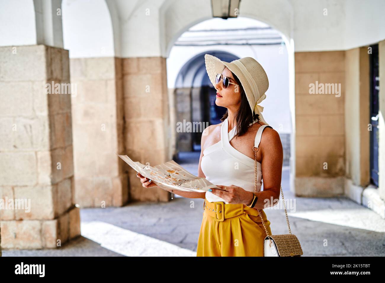 Thoughtful female tourist in hat and sunglasses reading paper map while ...