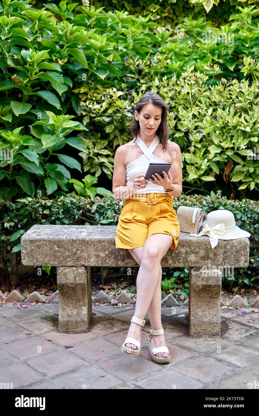 Full Body Of Tranquil Female Sitting On Stone Bench And Browsing Tablet