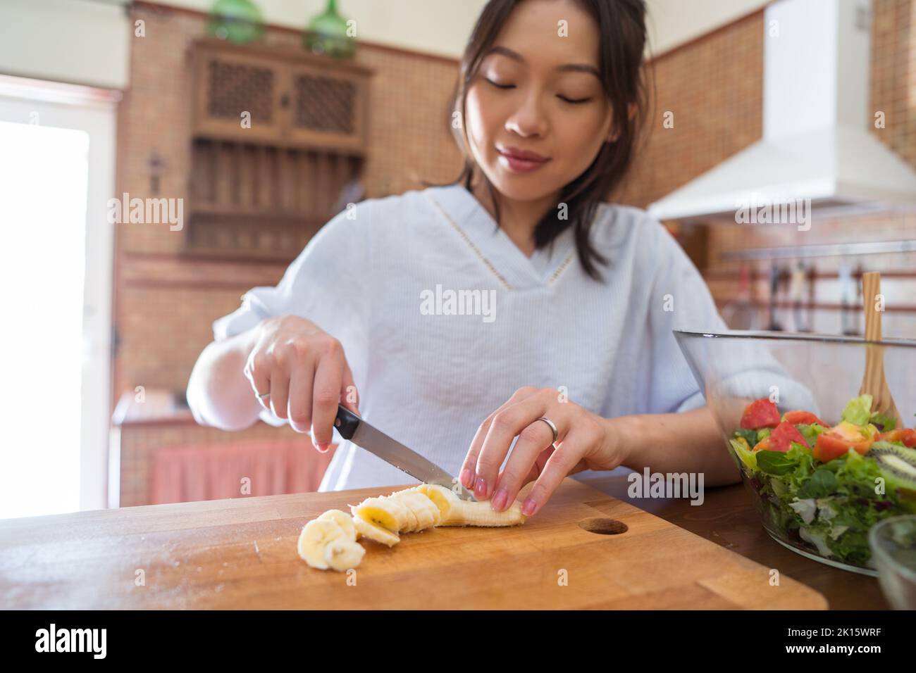 Young Asian female cutting fresh banana on chopping board near bowl ...