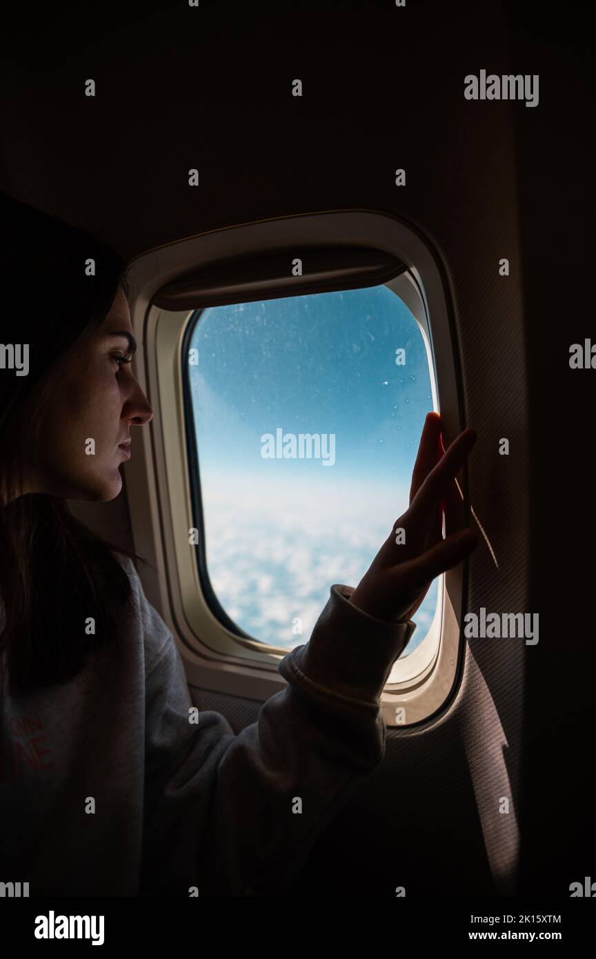 Thoughtful woman admiring cloudy blue sky through window of dark plane ...