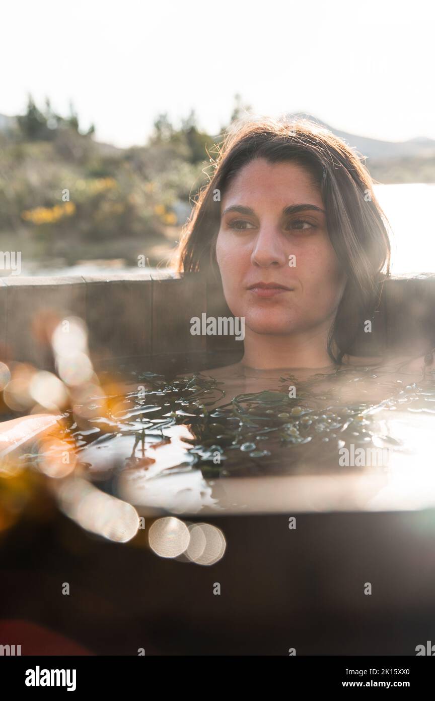 Thoughtful young female taking hot bath with seaweed in barrel on lake ...