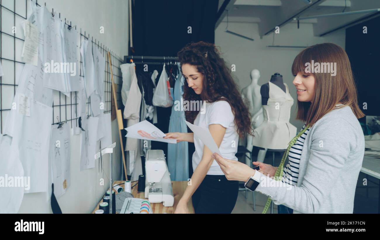 Attractive women clothing designers looking at garment sketches hanging ...