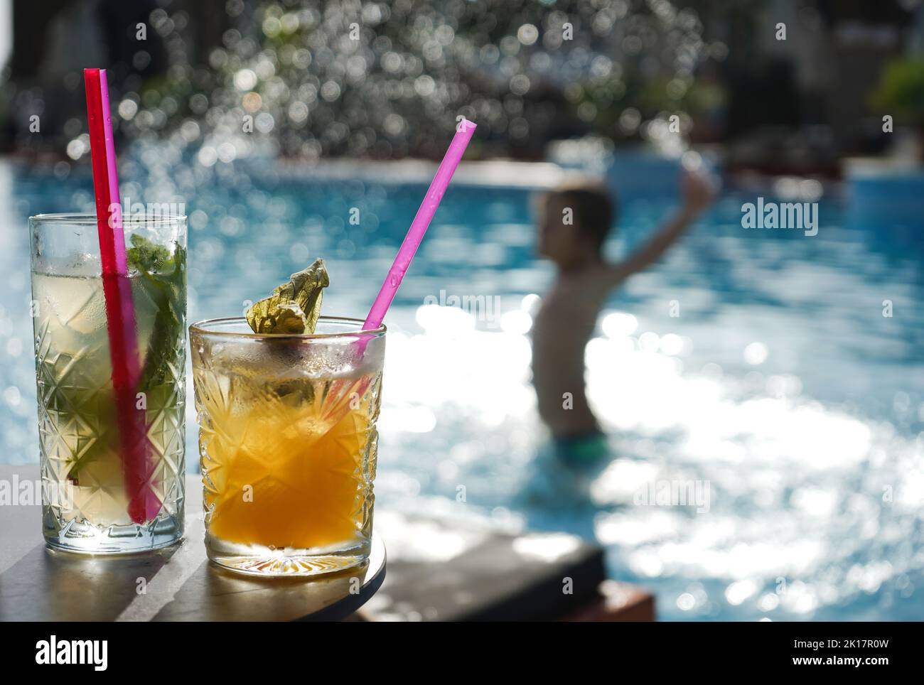 focus on two fruit cocktails standing poolside with kid playing in ...