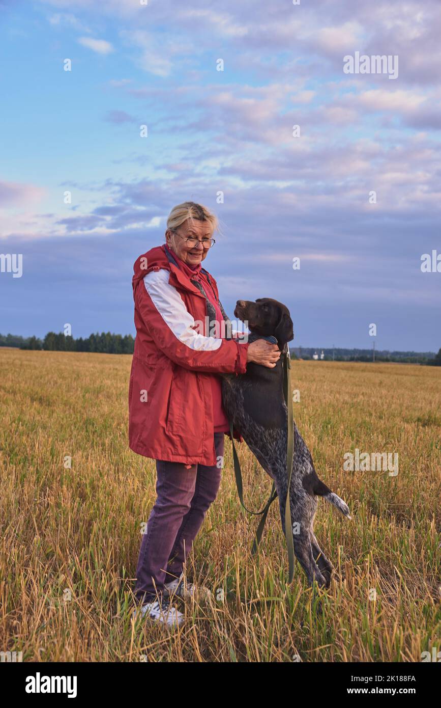 An elderly woman hugs her dog , German shorthaired pointer , in a mown field at sunset. Active ...