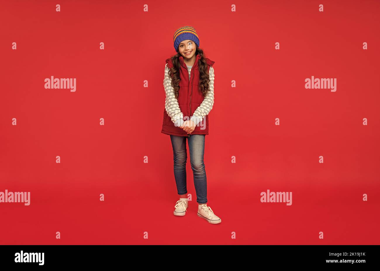 full length of smiling child in knitted winter hat and down vest on red ...