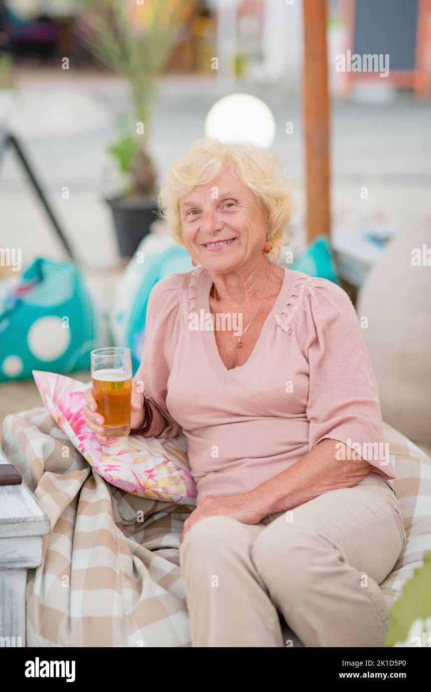 Mature woman sitting at a table in a summer cafe and drinking beer ...