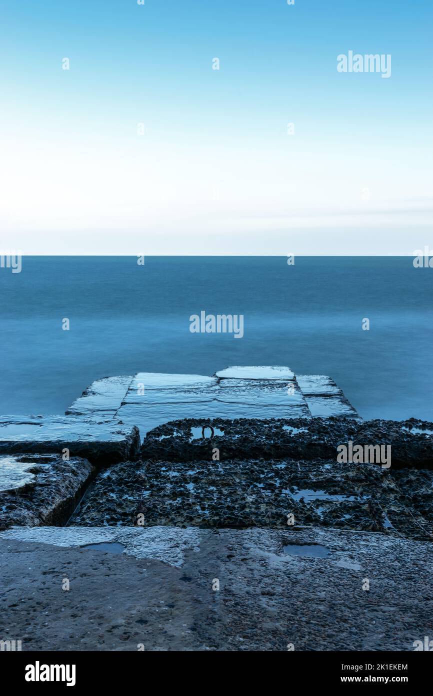 A vertical shot of a broken stone pier at lake shore under blue sky ...