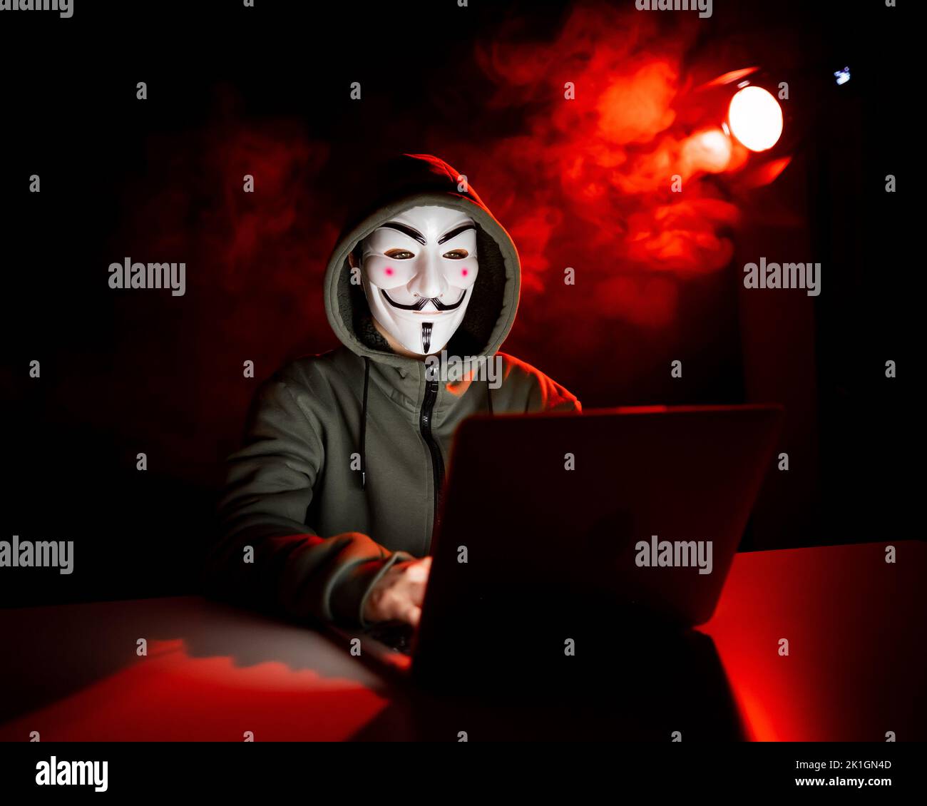Anonymous in a hood is typing on a laptop in the dark in red smoke ...