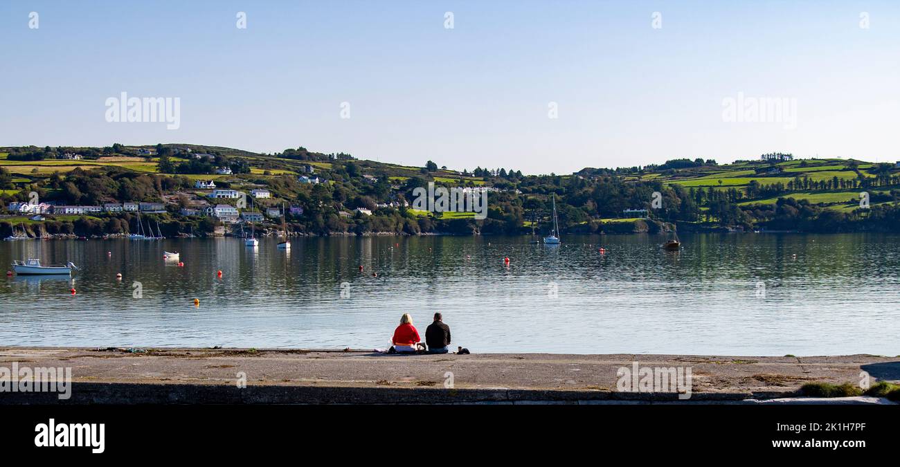 Couple man and woman sat on pier alone looking at Glandore Harbour ...
