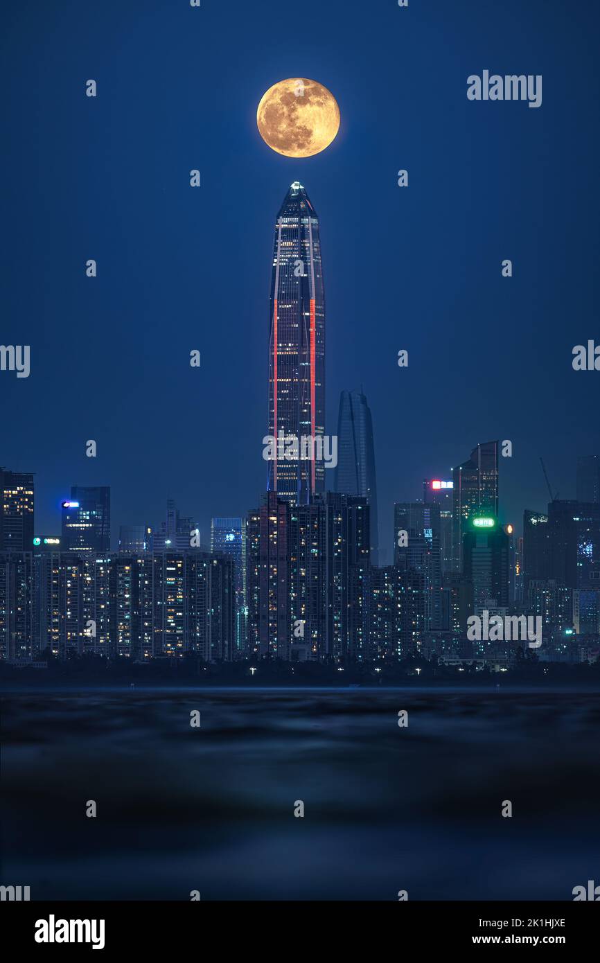 A vertical shot of full moon over a tower in a modern city's skyline at ...