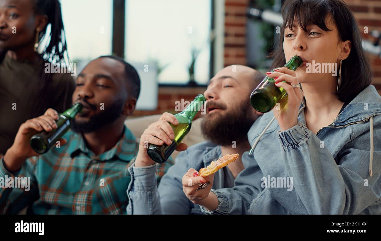 Multiethnic group of friends watching comedy movie on tv and clinking beer bottles at social ...