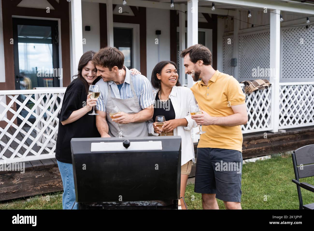 Smiling multiethnic couples with wine talking near grill in backyard ...