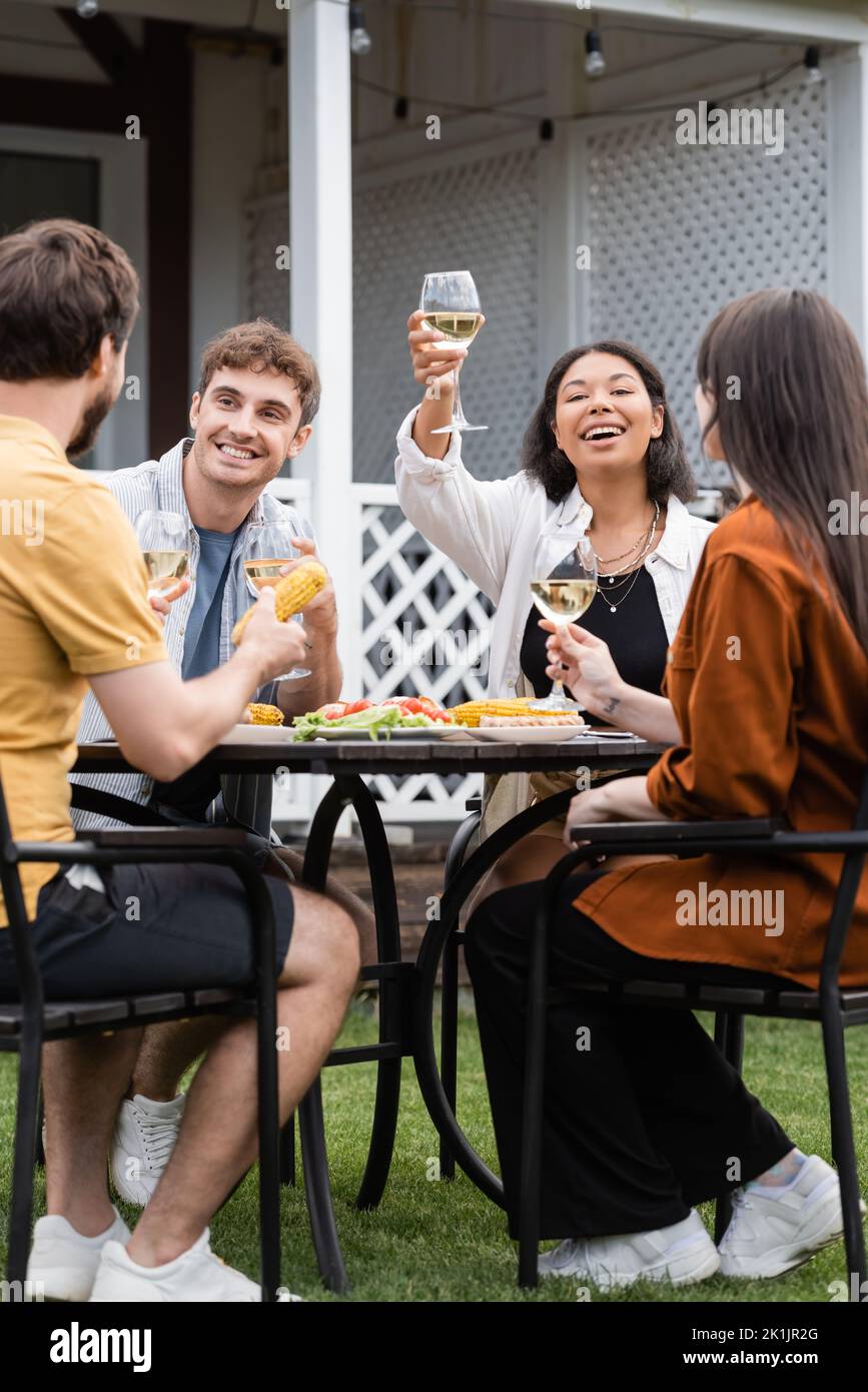 happy bi-racial woman raising glass of wine near friends during bbq ...