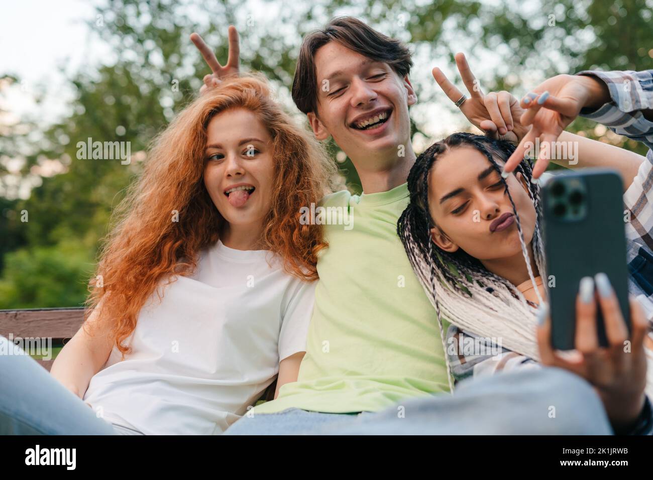 Three young smiling friends sitting on a park bench taking self ...