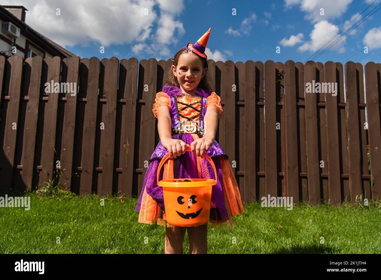 girl in clown costume and party cap standing with halloween bucket in ...