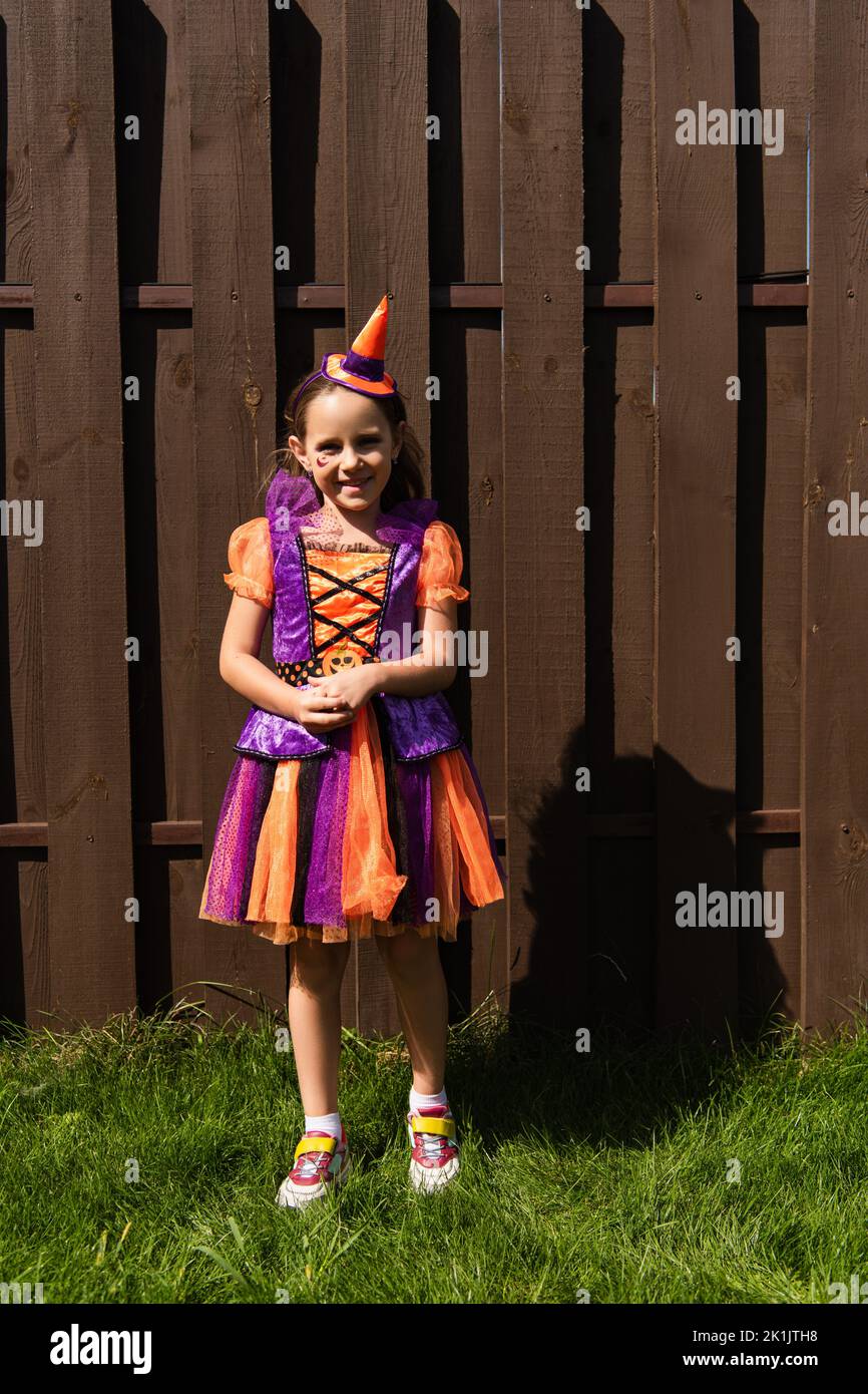 full length of girl in clown costume and party cap smiling near wooden ...