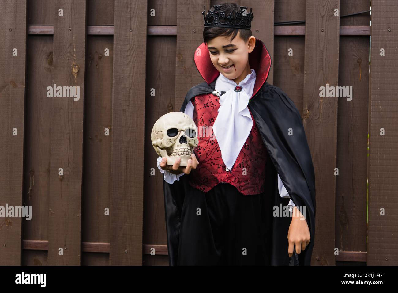happy asian boy in vampire king costume standing with spooky skull near ...