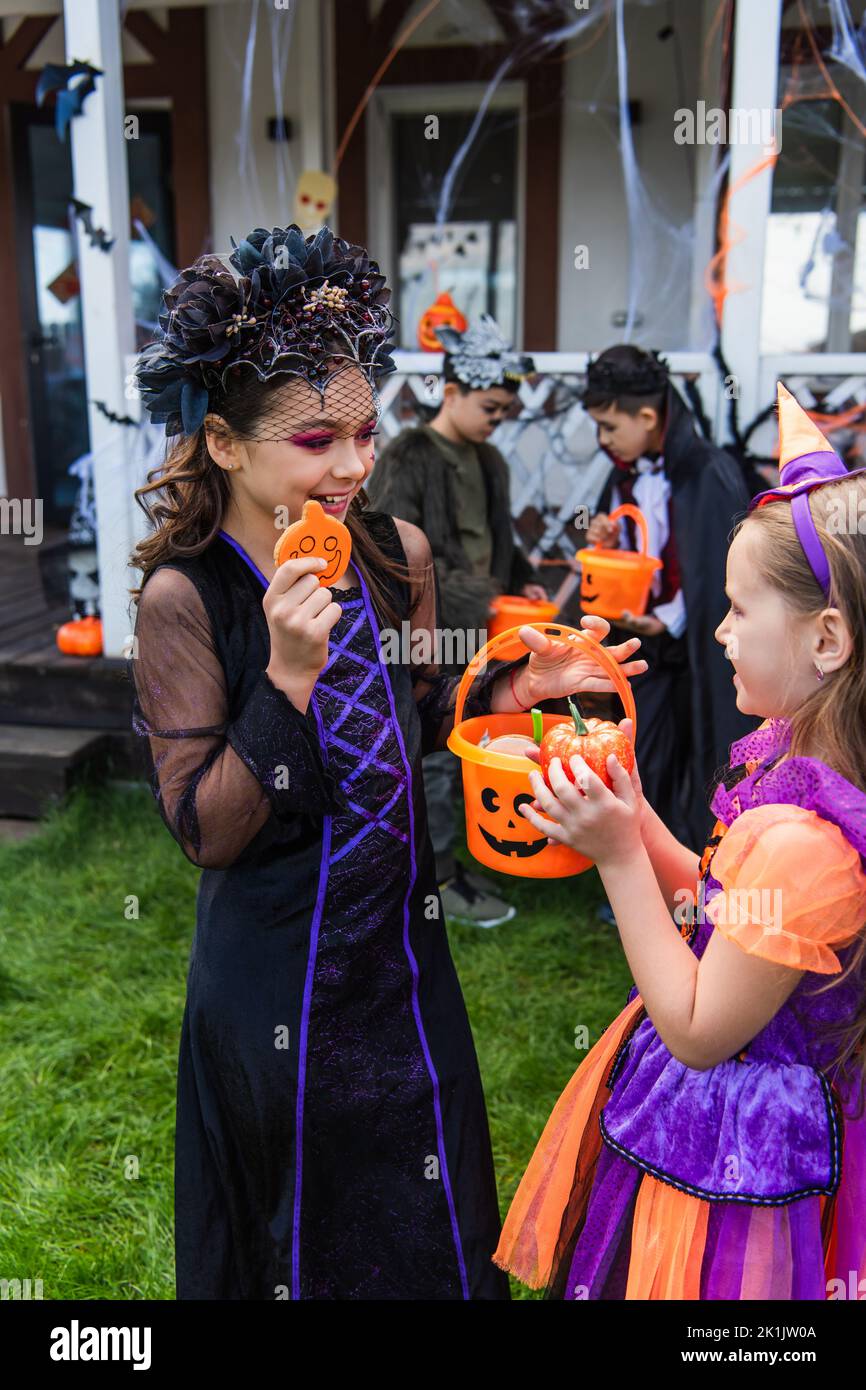 Smiling kid in halloween costume holding candy and bucket near friend ...