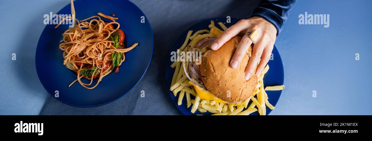 top view of cropped woman taking burger near french fries and spaghetti ...