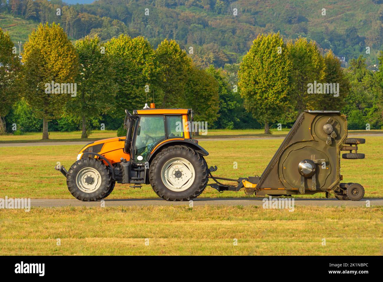 Yellow tractor with an industrial lawn mower trailer drives along the ...