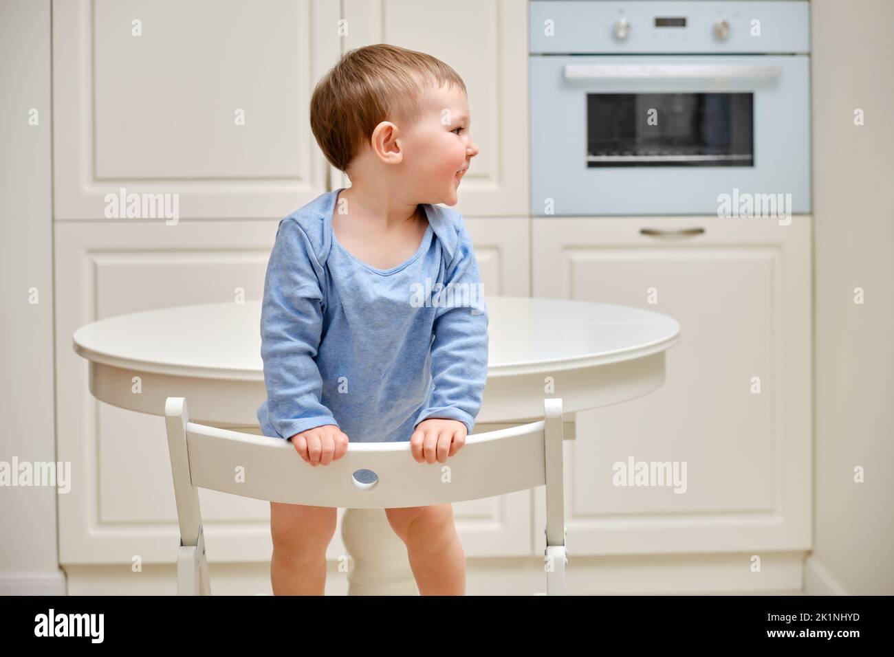Toddler baby climbs onto a chair at the kitchen white table. A child in ...