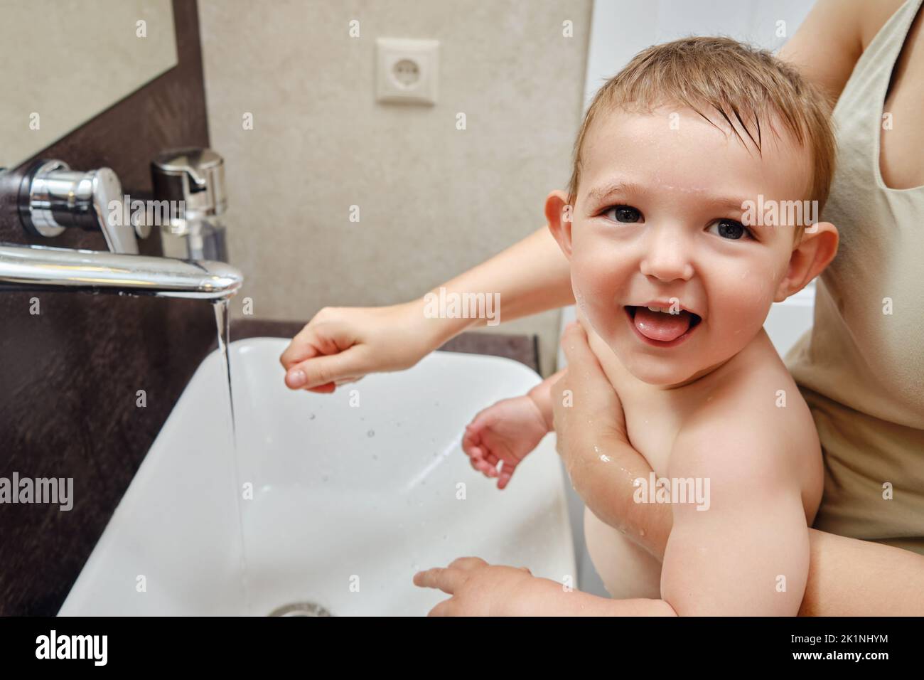 Woman mother washes face of happy toddler baby in sink with faucet. Mom ...