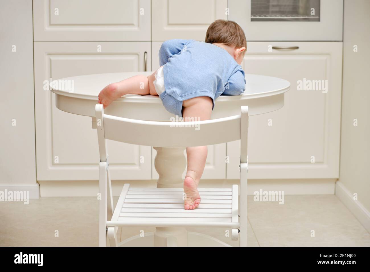 Toddler baby climbs on the kitchen white table. A child in danger ...