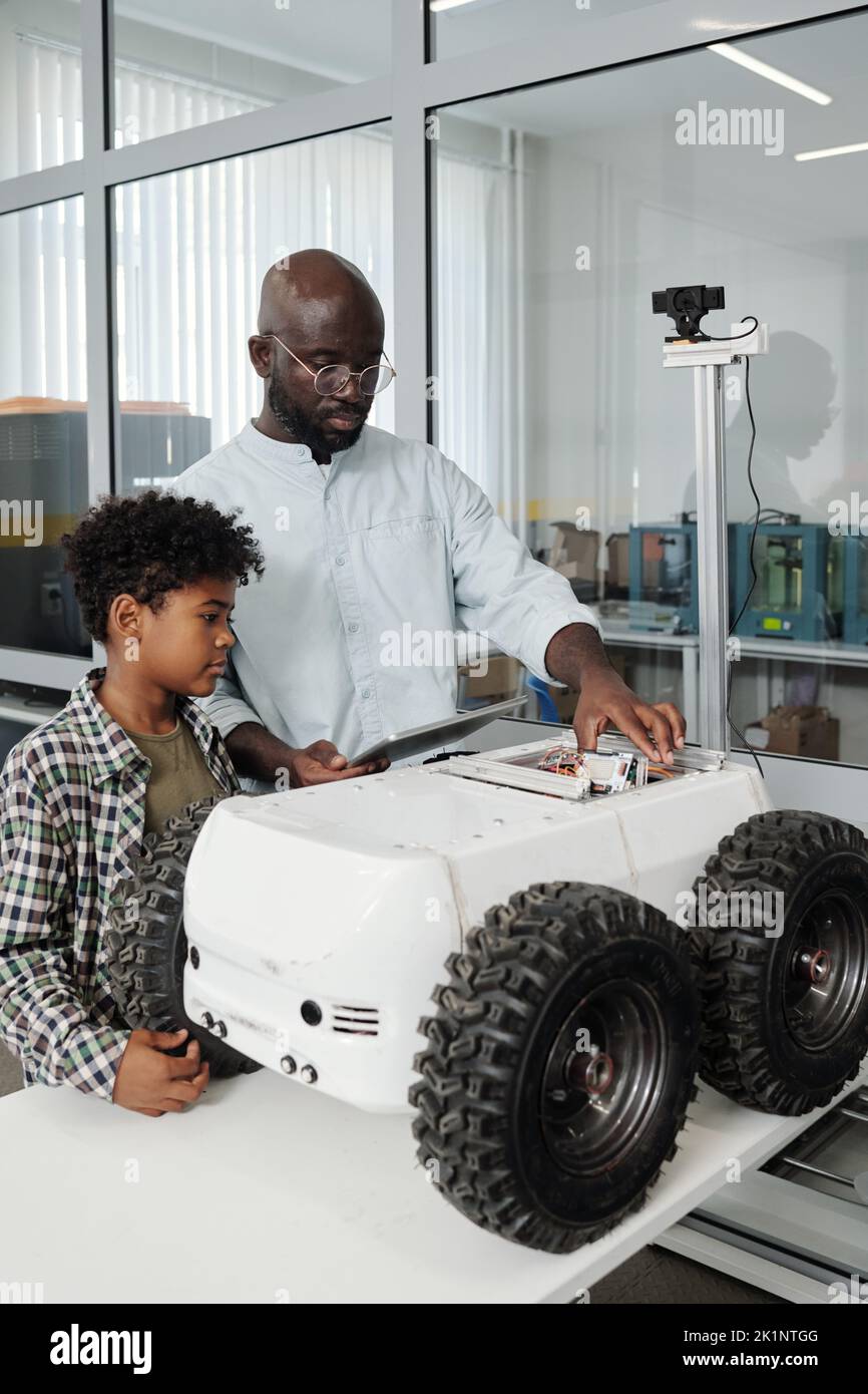 Young confident teacher with tablet explaining schoolboy how to control crawler which has been ...