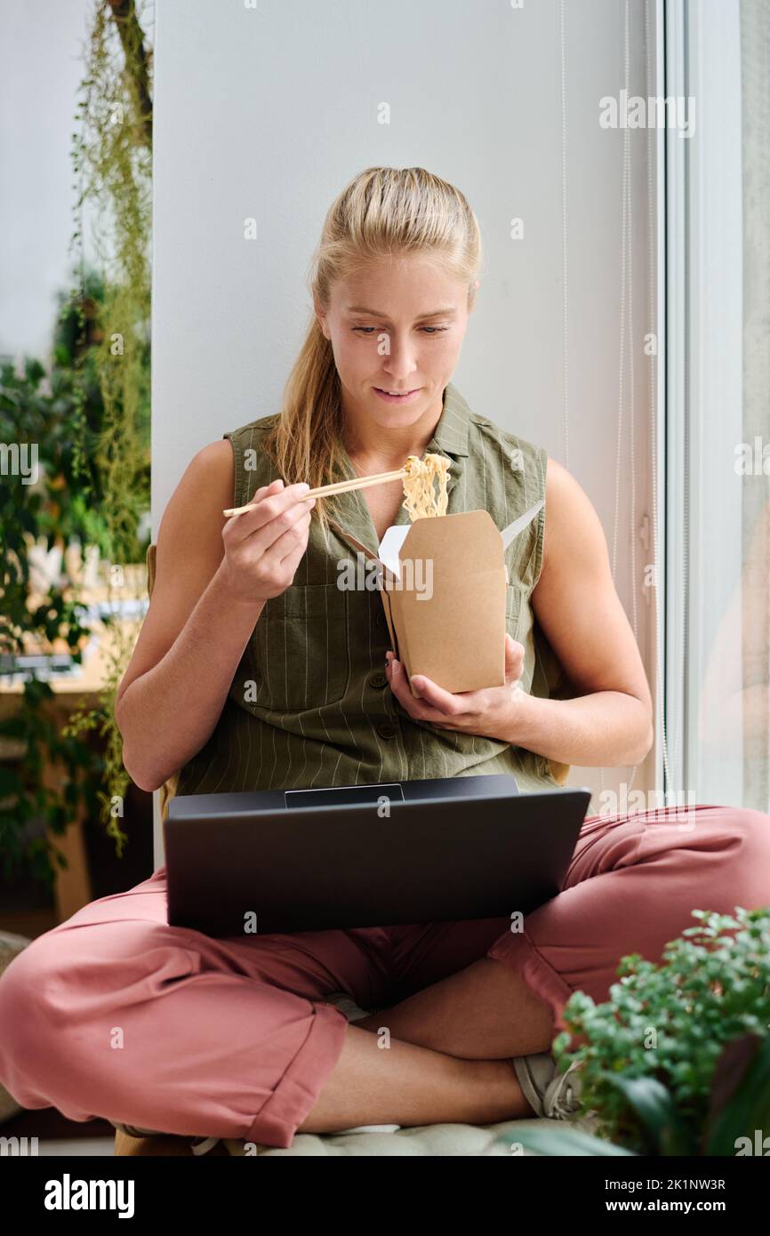 Young hungry businesswoman with laptop on her crossed legs eating ...