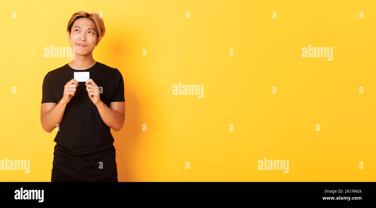 Thoughtful smiling asian guy thinking while showing credit card ...