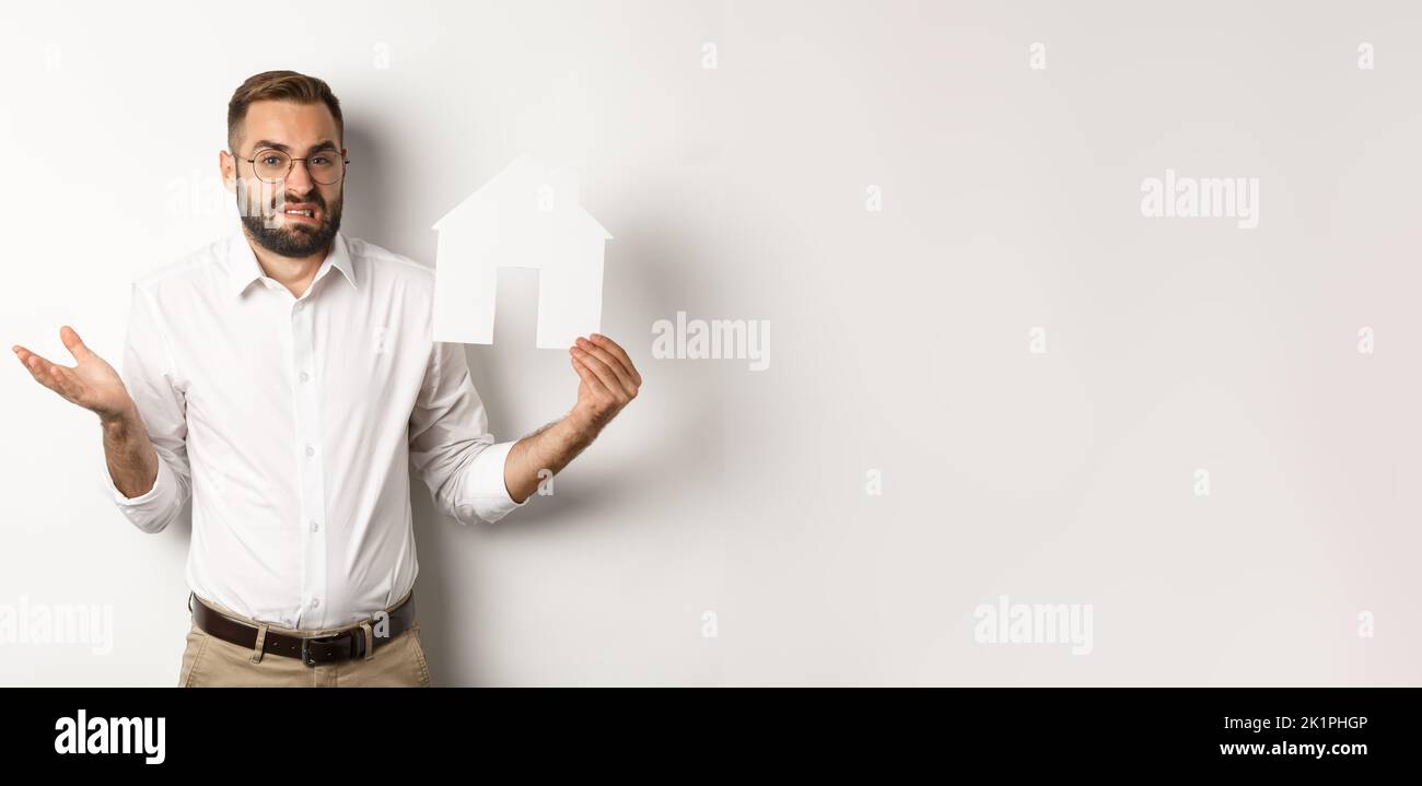 Real estate. Confused man shrugging, showing house paper model and ...