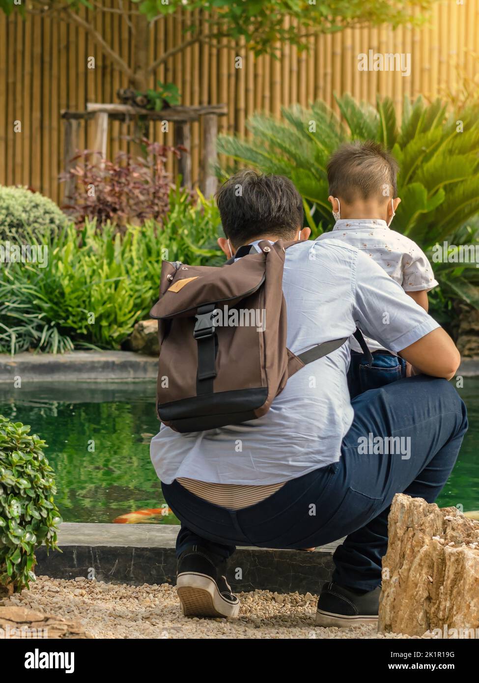 Back view of Asian father wear face mask with backpack embraces his son ...