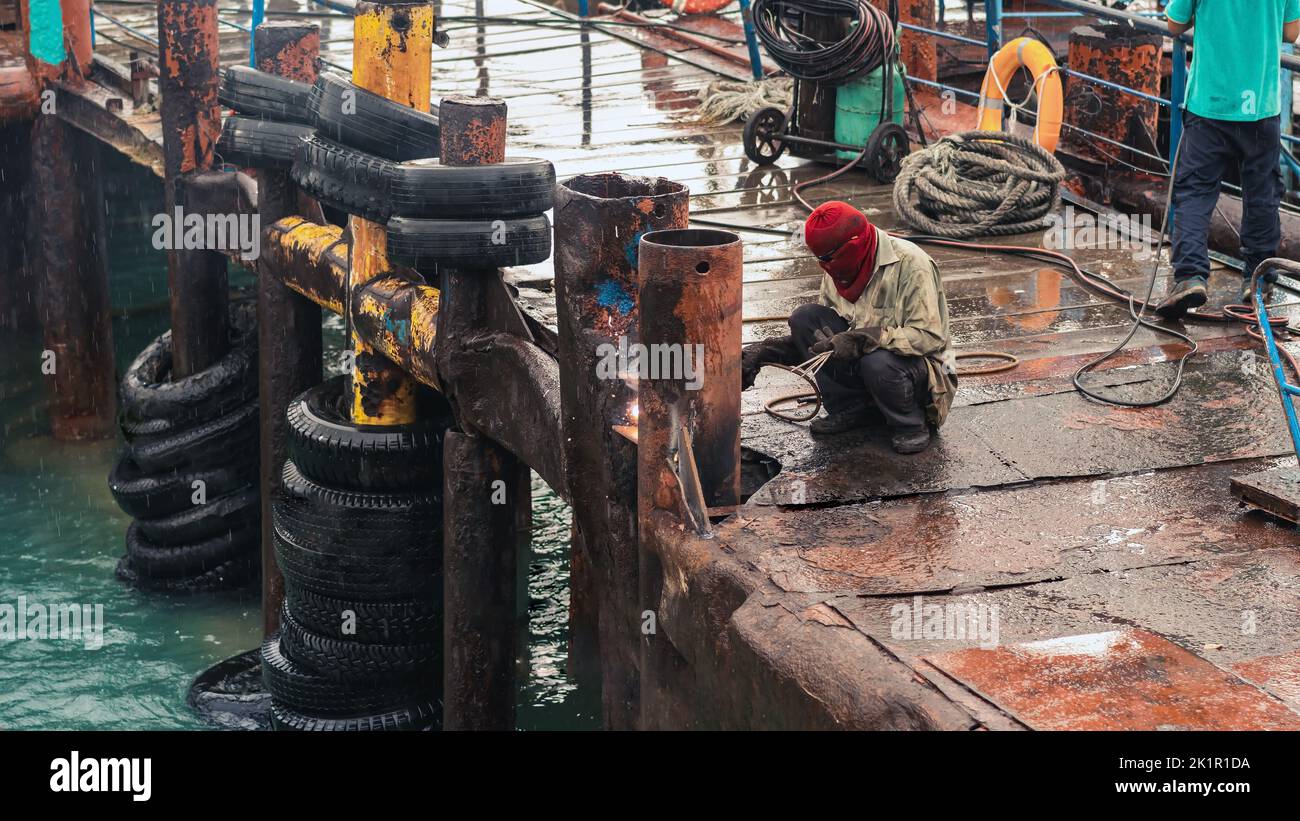 Worker welds steel parts by hand at the pier in the rain. Industrial ...