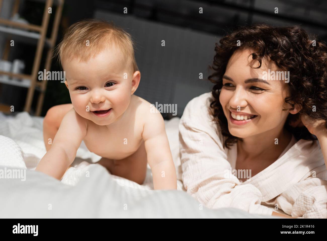 cheerful mother with curly hair looking at happy baby daughter crawling ...