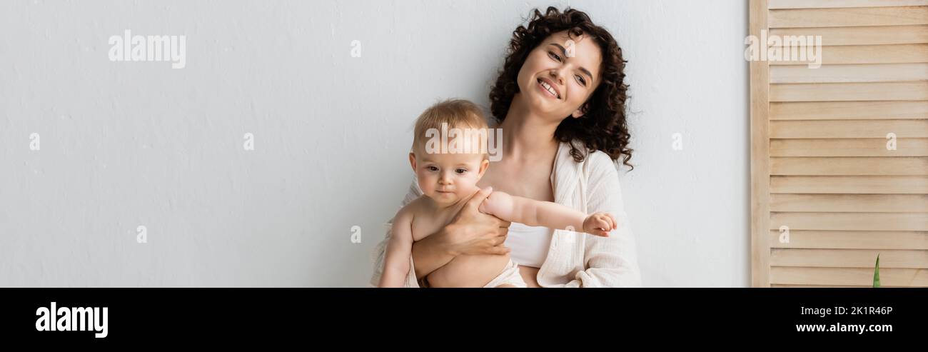 Smiling mother holding baby girl near folding screen at home, banner ...