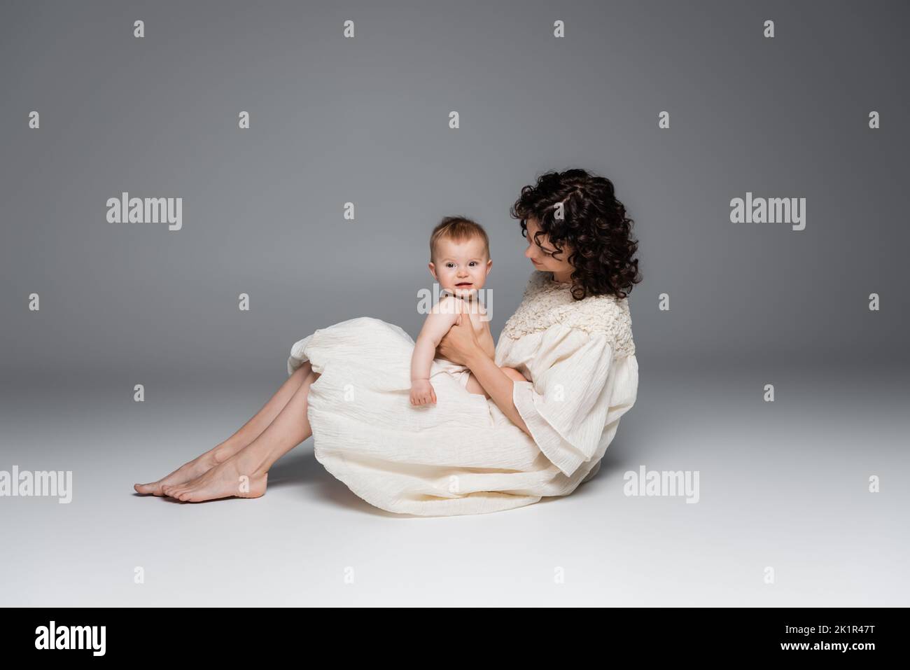 Curly mother in dress holding smiling bay girl while sitting on grey ...