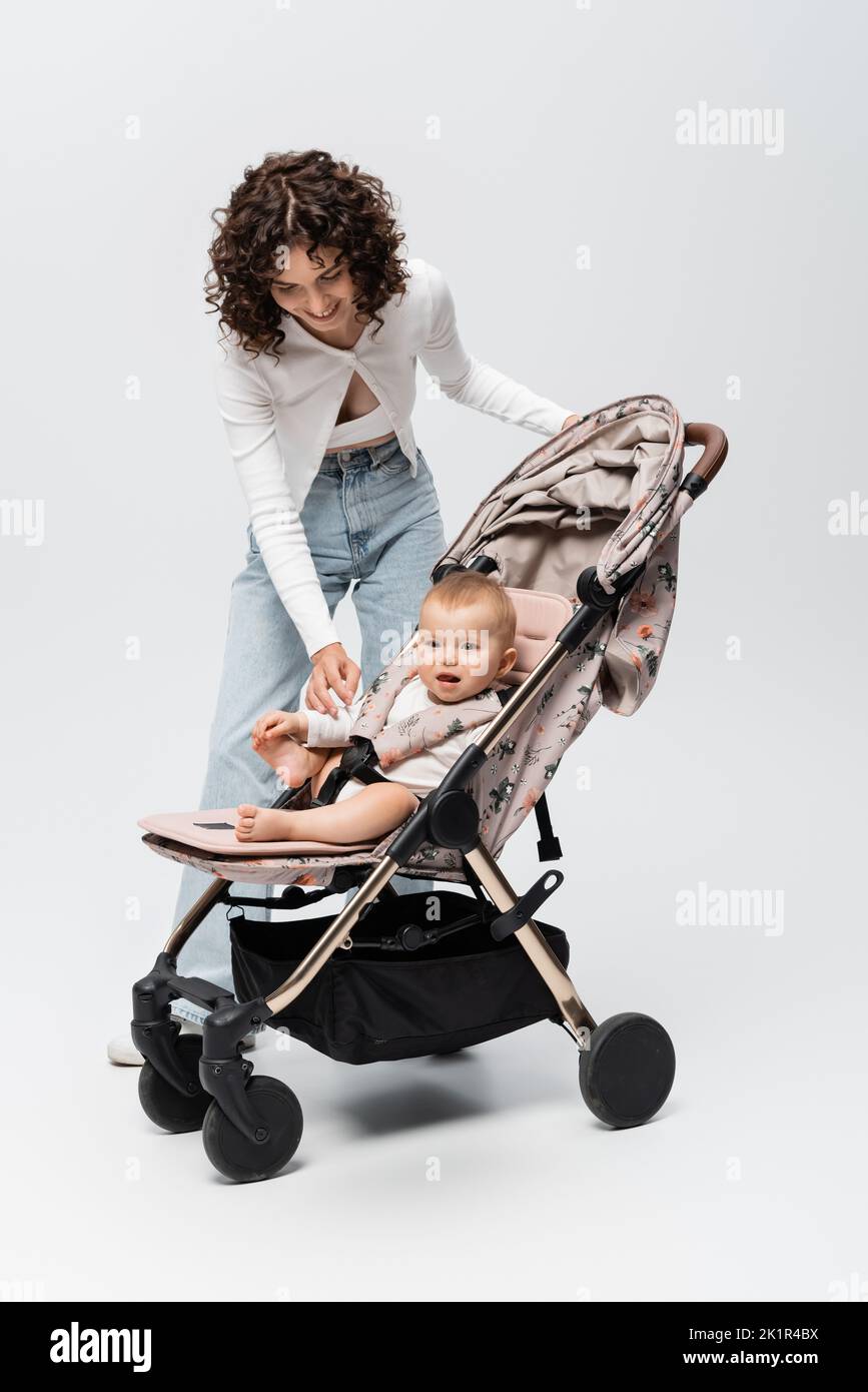Smiling woman looking at baby daughter in stroller on grey background ...