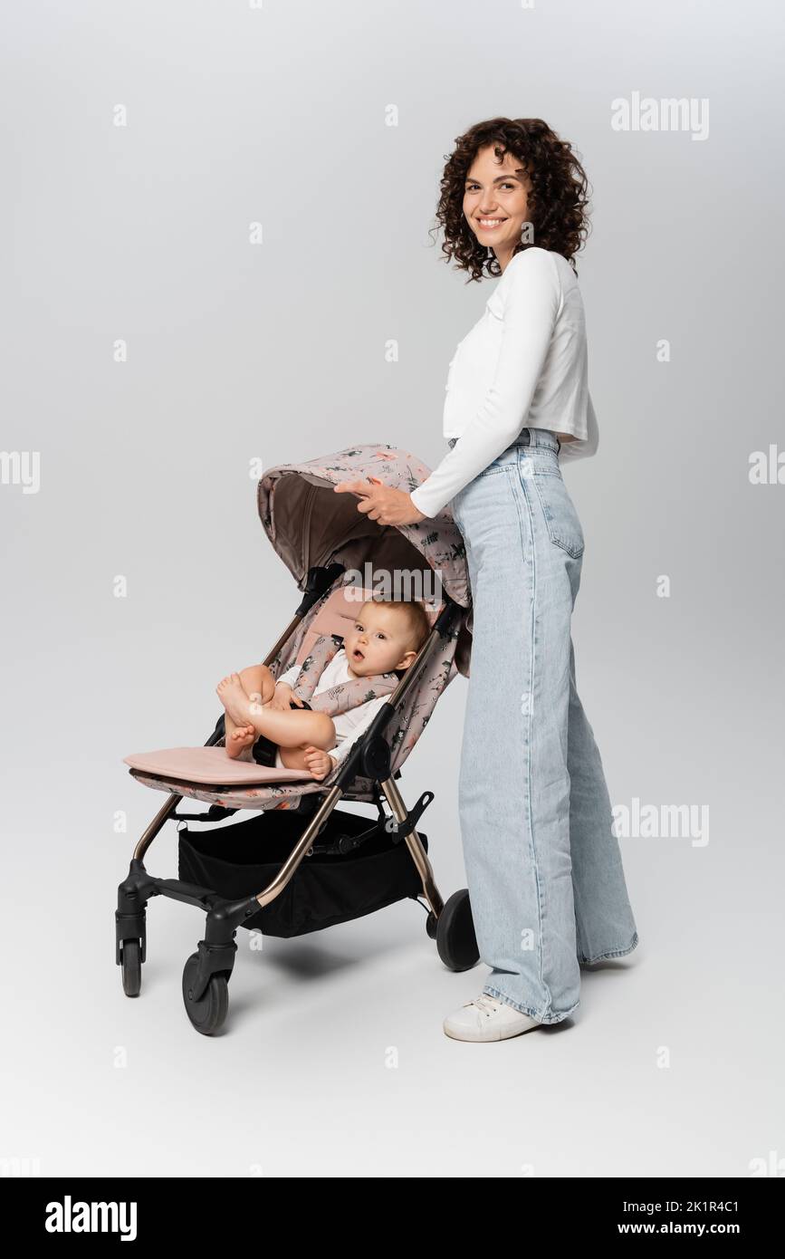 Curly mother in jeans standing near baby girl in stroller on grey ...