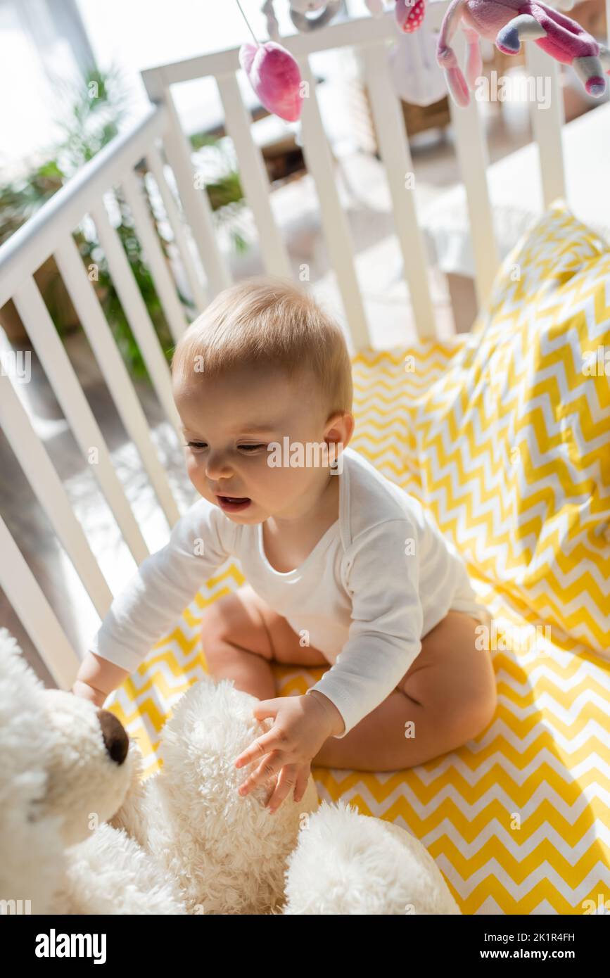 high angle view of cute infant girl in baby romper sitting near teddy ...
