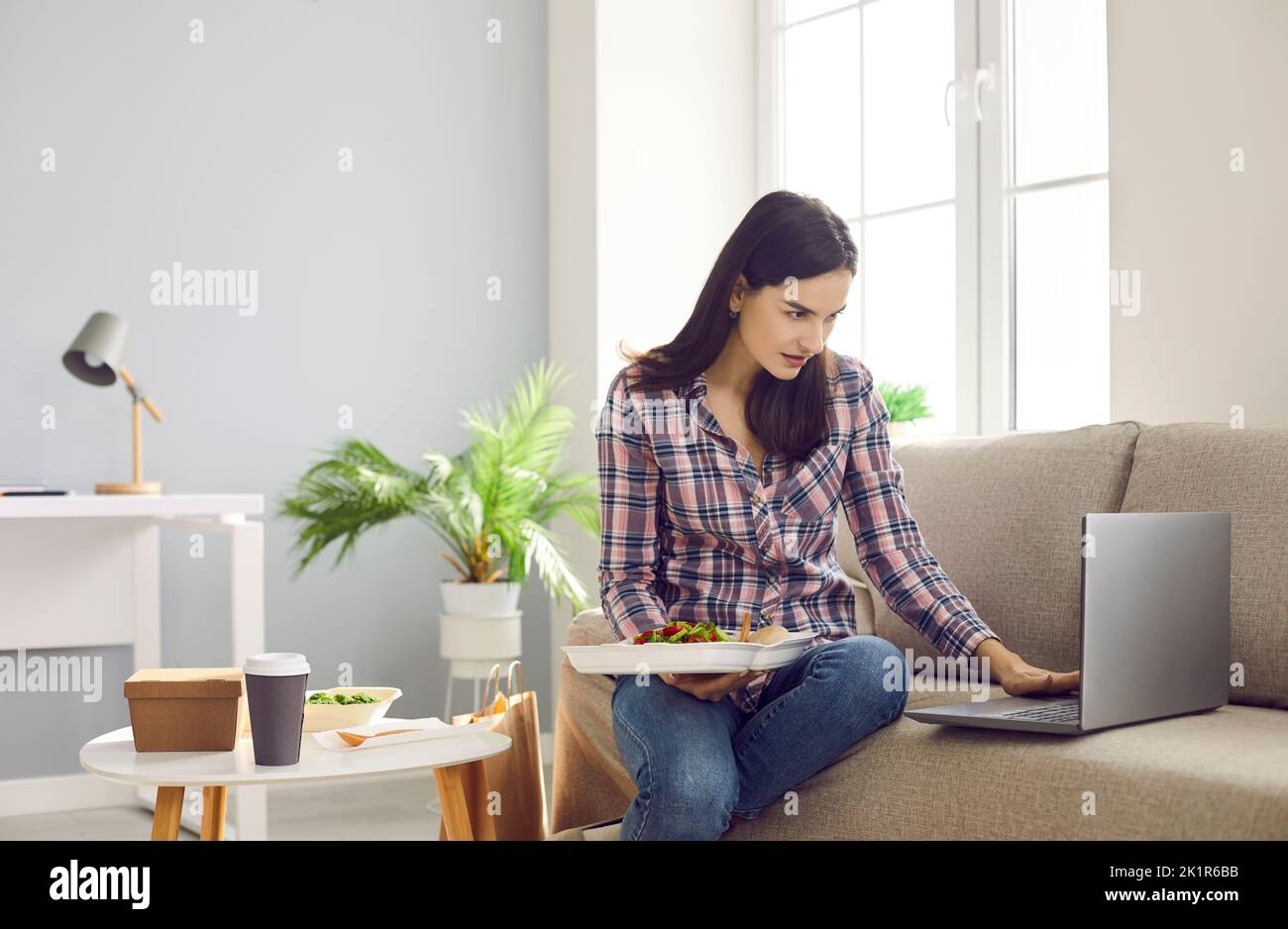 Busy young woman using laptop computer while eating takeaway meal on ...