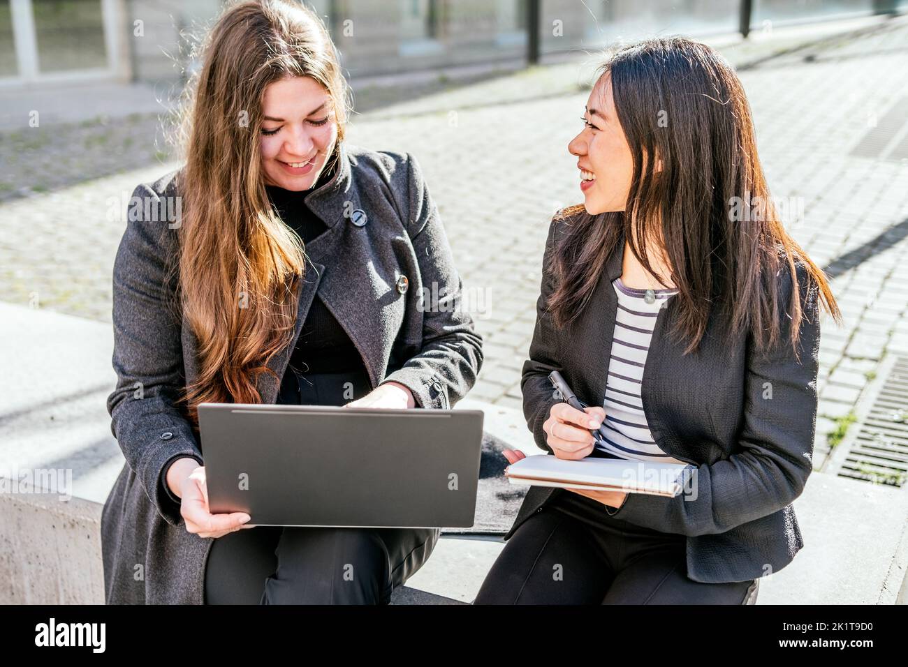 Positive woman typing on laptop while sitting near Asian female ...