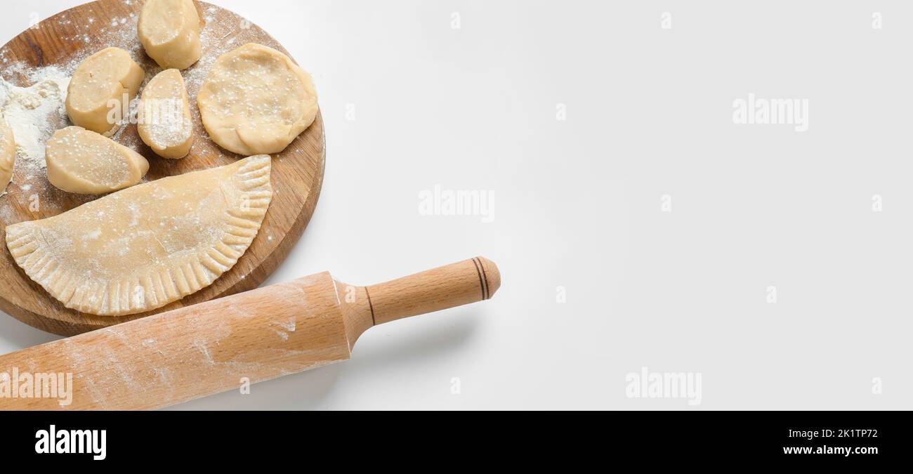 Board with raw cheburek, pieces of dough and rolling pin on white ...